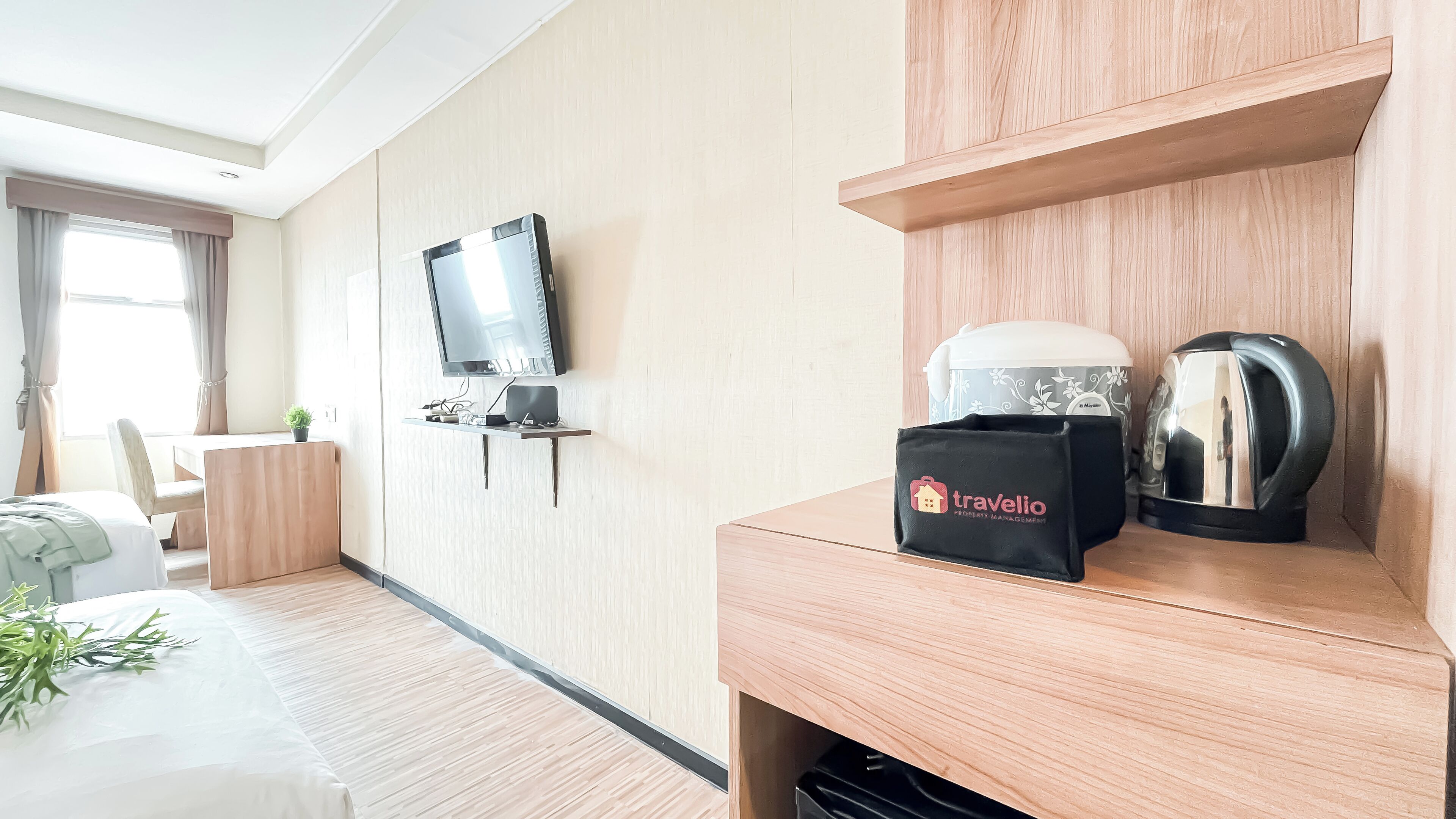 Quarto de hotel moderno com televisão de ecrã plano na parede, máquina de café, mala de viagem e uma pequena mesa com uma planta.