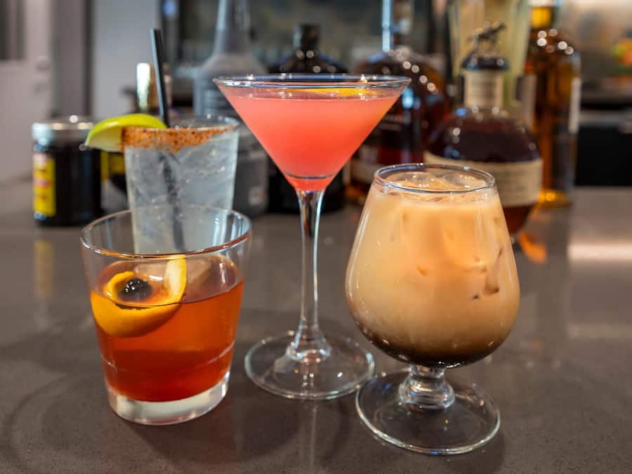 Três bebidas diferentes numa bancada de bar, com garrafas ao fundo.
