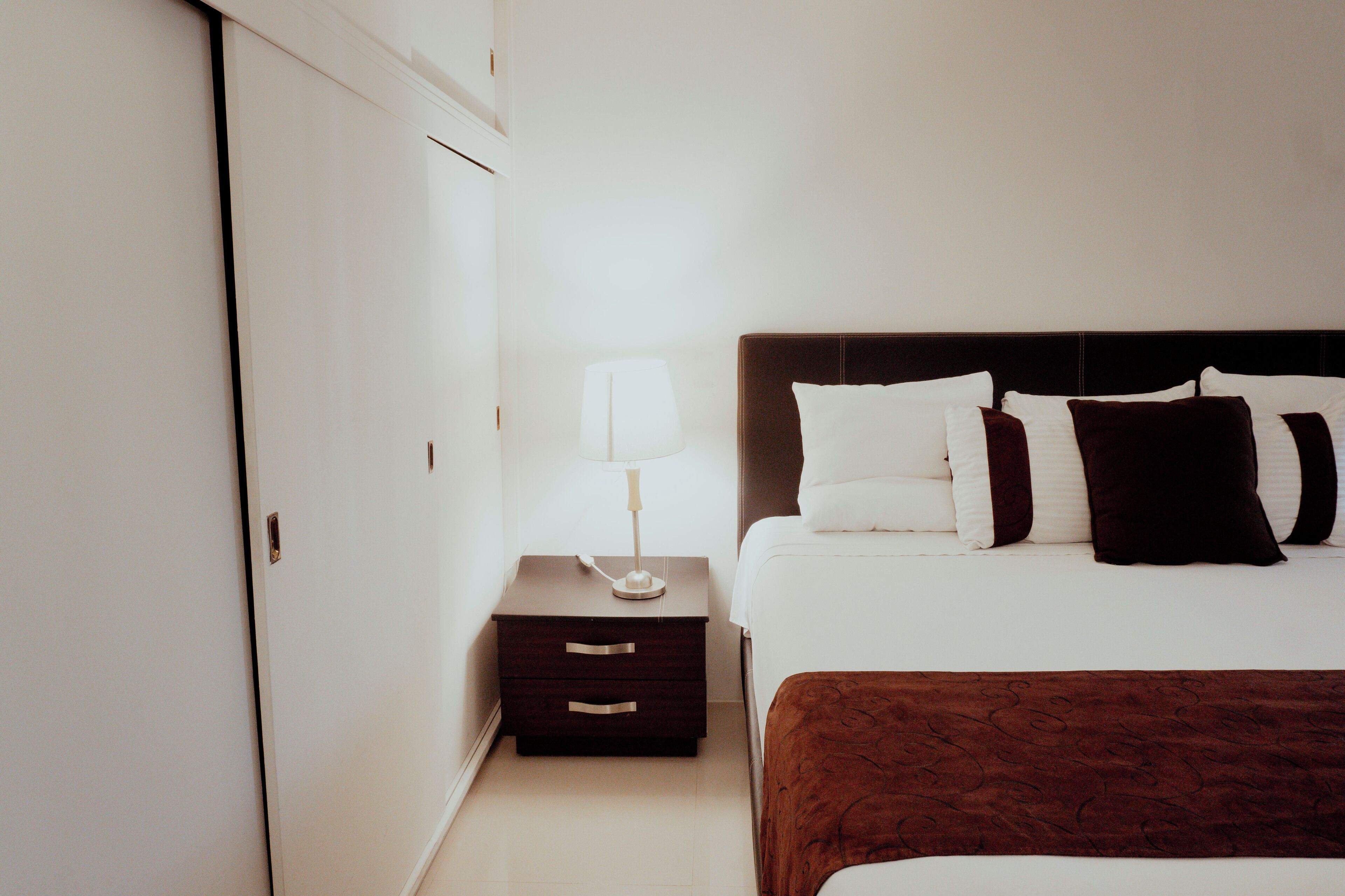 Quarto de hotel com cama, mesa de cabeceira com abajur e guarda-roupa.