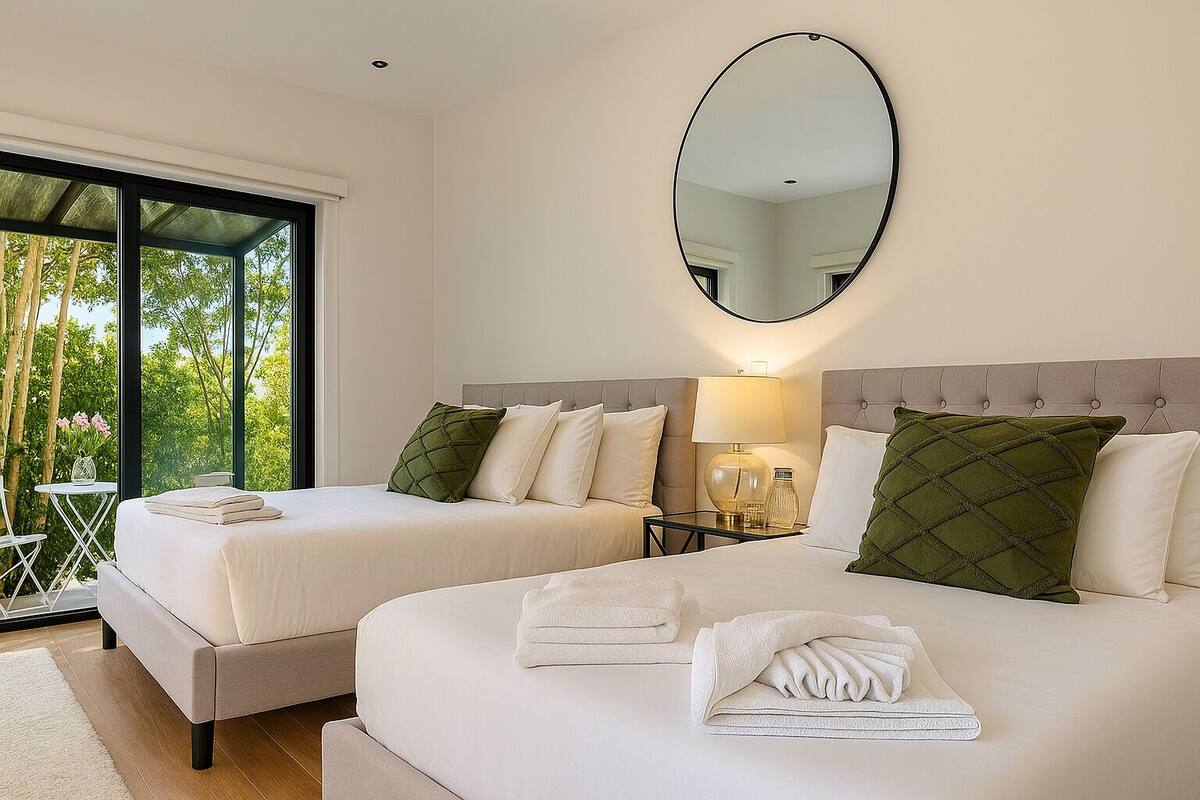 Quarto de hotel com duas camas, um espelho grande, mesa de cabeceira com abajur e vista para o verde pela janela.