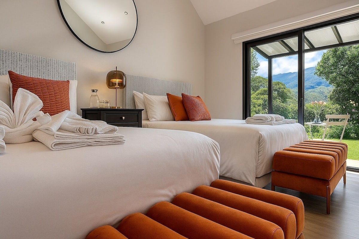 Quarto de hotel moderno com duas camas, uma janela grande com vista para as montanhas e uma pequena mesa com abajur.