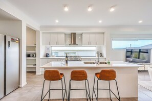 Uma cozinha moderna com armários brancos, uma ilha central com banquetas, um exaustor de aço inoxidável e uma grande janela com vista para o exterior.