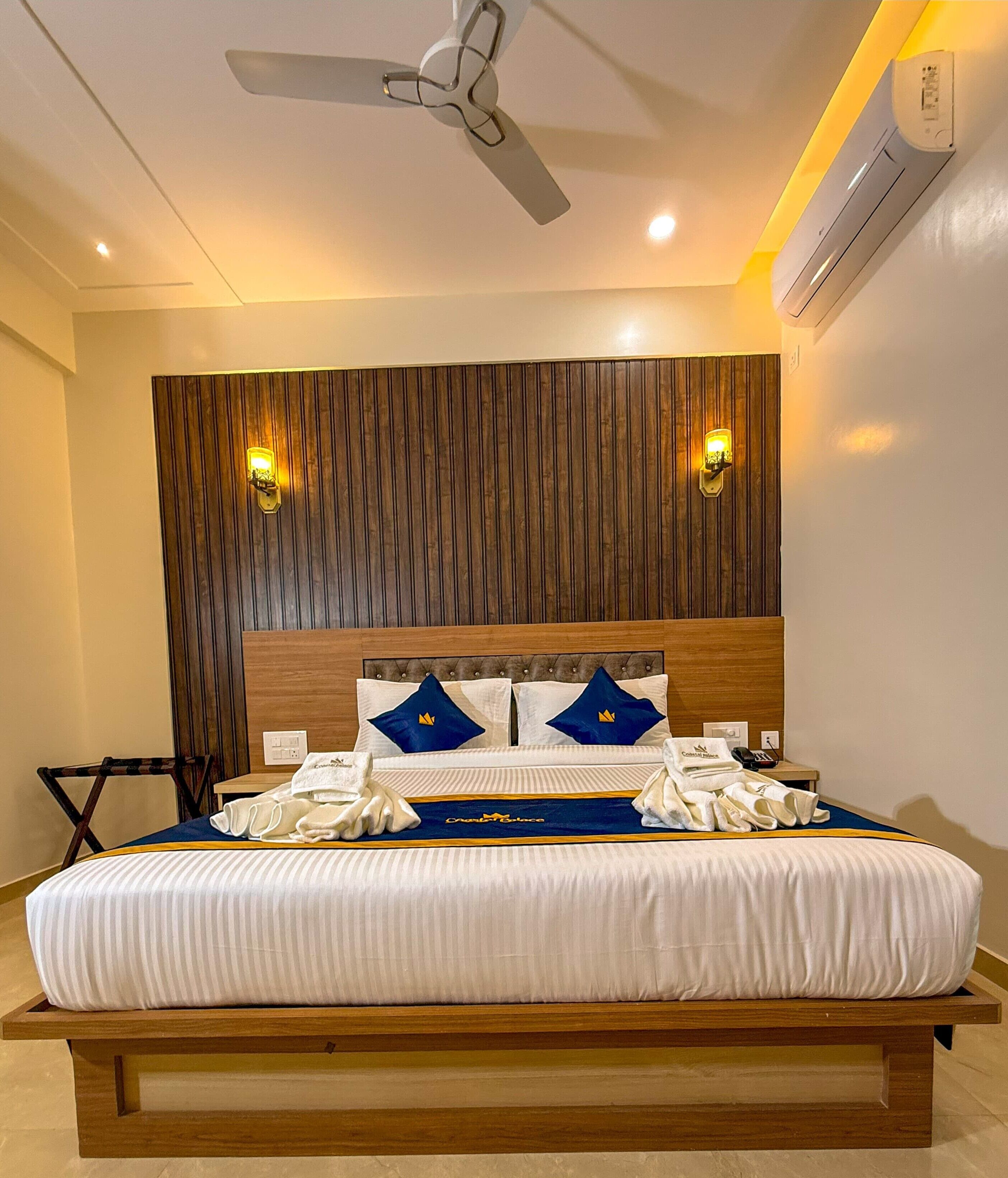 Uma cama bem arrumada com cabeceira de madeira, duas luminárias na parede e um ventilador de teto.