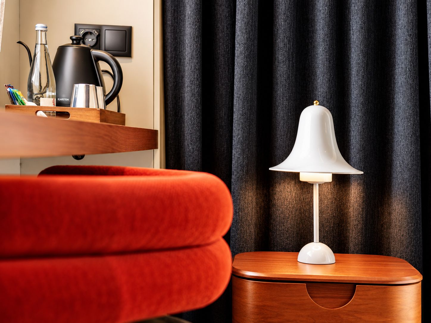 Quarto de hotel moderno com mesa de cabeceira de madeira, luminária branca, cadeira vermelha, chaleira preta e cafeteira em mesa de madeira.