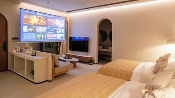 Quarto de hotel moderno com uma cama grande, uma TV, área de estar com mesa de centro e uma tela na parede exibindo opções de filmes.