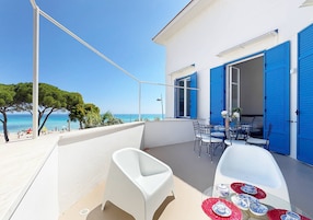 Varanda com cadeira branca, mesa com pratos e persianas azuis numa construção com vista para a praia.