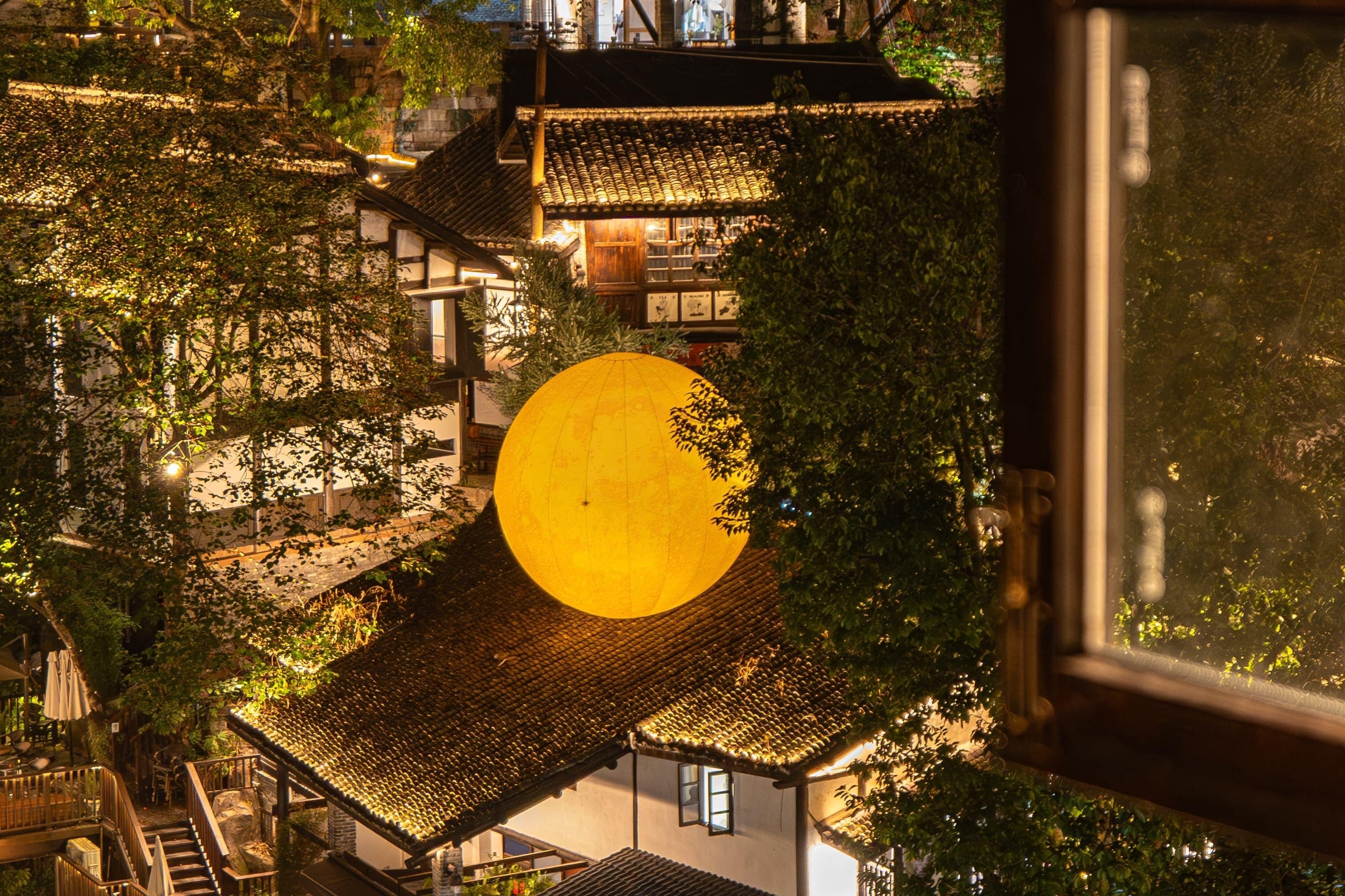 Um edifício japonês tradicional com uma grande estrutura iluminada em forma de lua no centro, rodeado por árvores e arquitetura tradicional.