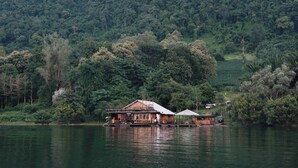 Uma casa flutuante de madeira num lago, rodeada por floresta densa.