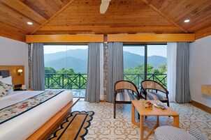 Quarto de hotel com teto de madeira, uma cama grande com colcha florida, duas cadeiras, uma mesa pequena e vista para montanhas através de janelas amplas.