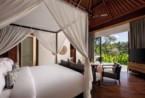 Quarto com cama grande, televisão, área de refeições com mesa e cadeiras, e vista para o verde lá fora.