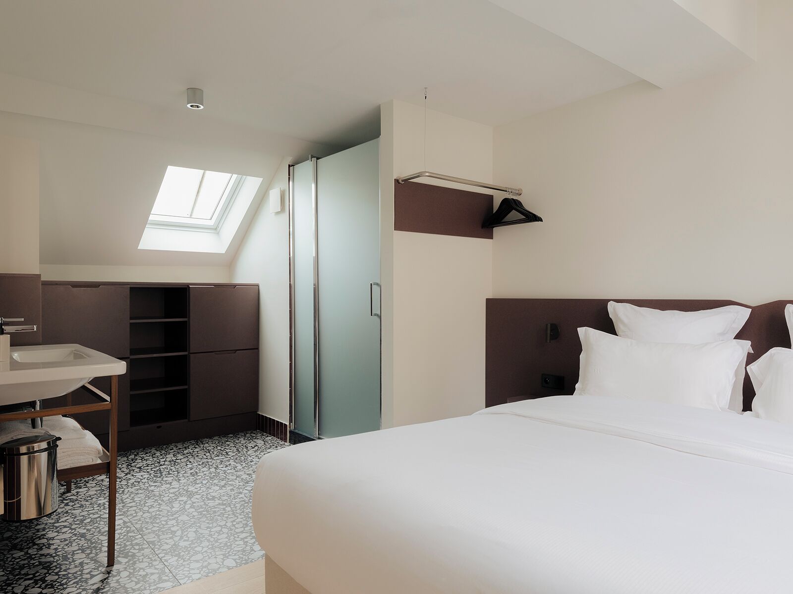 Quarto de hotel com cama, escrivaninha, espelho, guarda-roupa e claraboia.