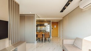Quarto de hotel moderno com televisão de tela plana, um sofá, área de refeições com cadeiras e uma cozinha visível através de um painel de vidro.