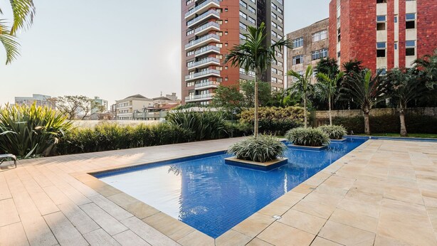 Piscina com deck revestido, rodeada por vegetação e edifícios altos ao fundo.