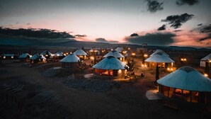 Diversas yurts com telhados cônicos, dispostas em círculo, iluminadas por luzes quentes sob um céu crepuscular.