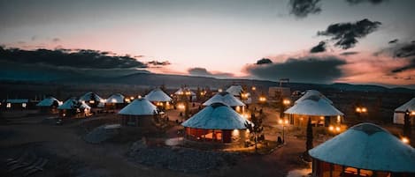 Diversas yurts com telhados cônicos, dispostas em círculo, iluminadas por luzes quentes sob um céu crepuscular.