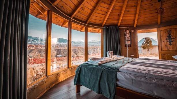 Quarto com teto de madeira alto, janelas amplas, uma cama e vista para uma paisagem montanhosa.