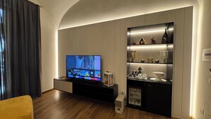 Uma sala de estar moderna com uma TV de tela plana em um suporte preto, piso de madeira, uma poltrona amarela e uma estante embutida com objetos decorativos.