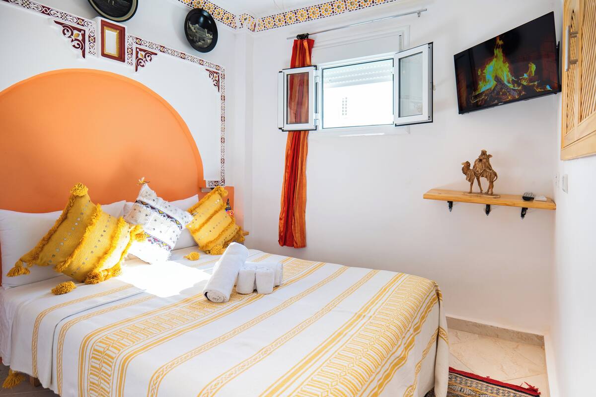 Quarto de hotel com uma parede laranja grande, cama com roupa de cama listrada, televisão montada e uma pequena prateleira com um objeto decorativo.