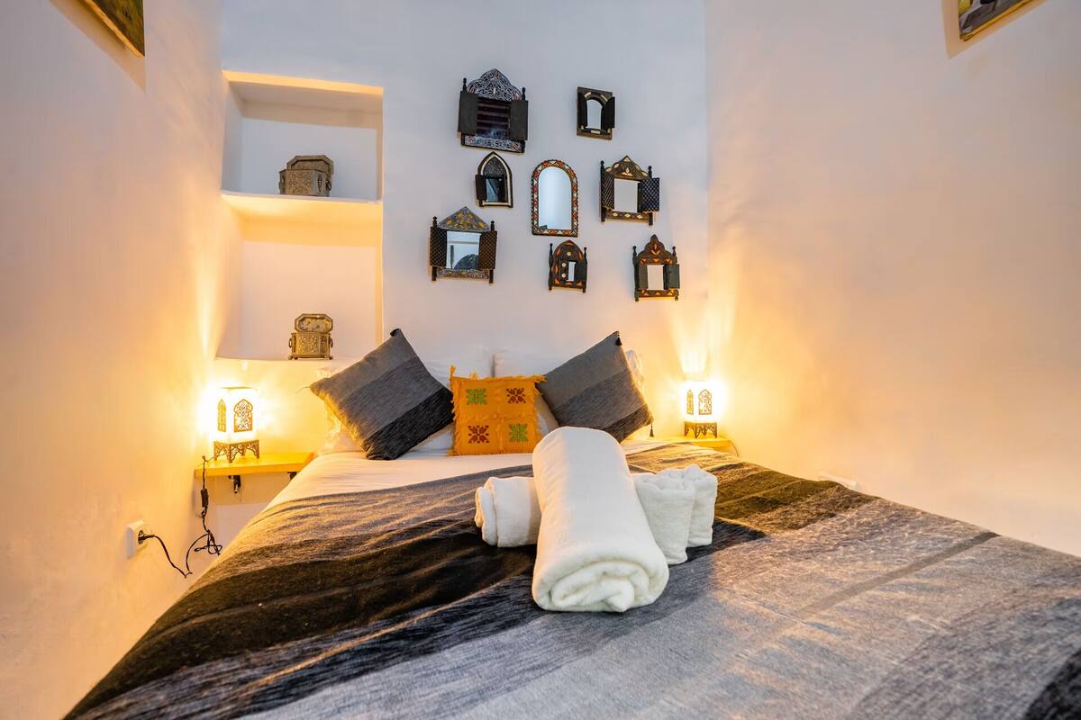 Cama bem arrumada com colcha estampada, duas almofadas e toalhas dobradas. Há luminárias fixadas na parede e espelhos decorativos sobre a cama.