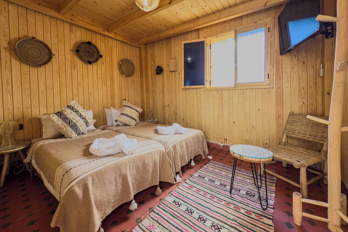 Quarto de madeira com duas camas, uma mesa redonda pequena e uma cadeira de fibra natural.