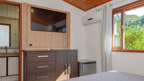 Quarto com cama, guarda-roupa de madeira, pequeno frigorífico, televisão na parede e janela com vista para o verde.