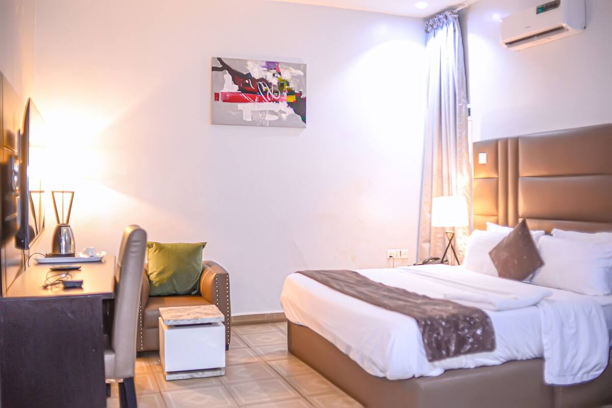 Quarto de hotel com uma cama, uma escrivaninha com cadeira, uma mesinha, uma luminária e um ar-condicionado fixado na parede.