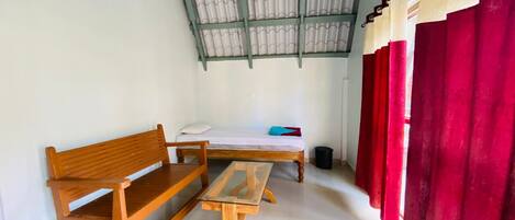 Um quarto com um banco de madeira, uma mesa pequena, uma cama com lençol branco e uma cortina vermelha.