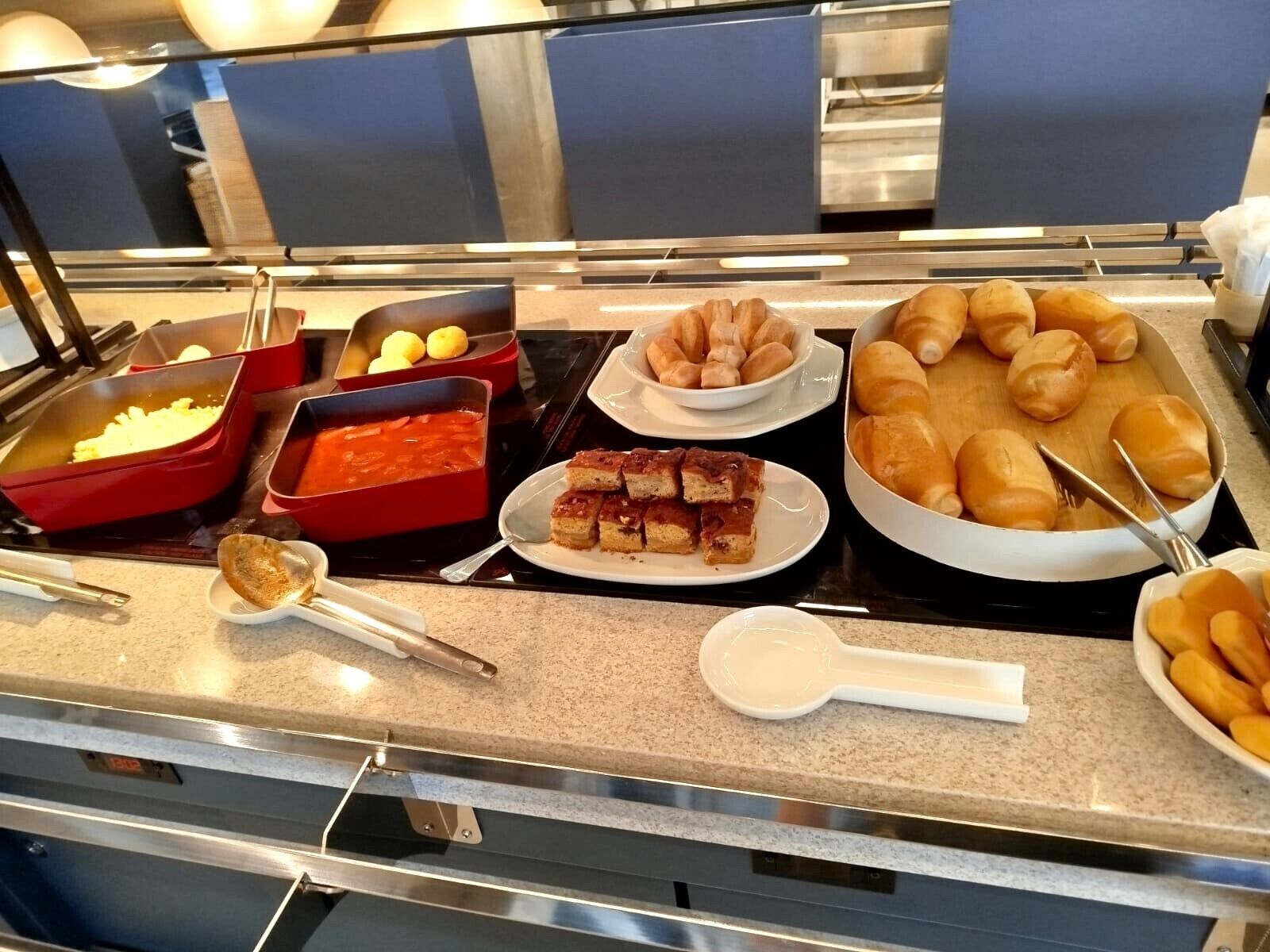 Buffet com diversos alimentos, incluindo pão, doces e um prato vermelho com uma colher branca.