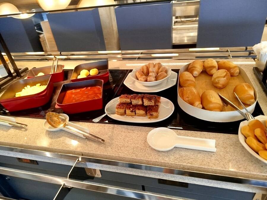 Buffet com diversos alimentos, incluindo pão, doces e um prato vermelho com uma colher branca.