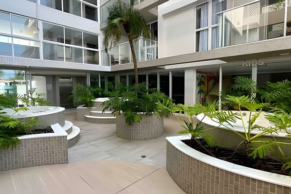 Um edifício moderno com vários andares, grandes janelas e pátios externos com vasos de plantas e um espelho d'água.