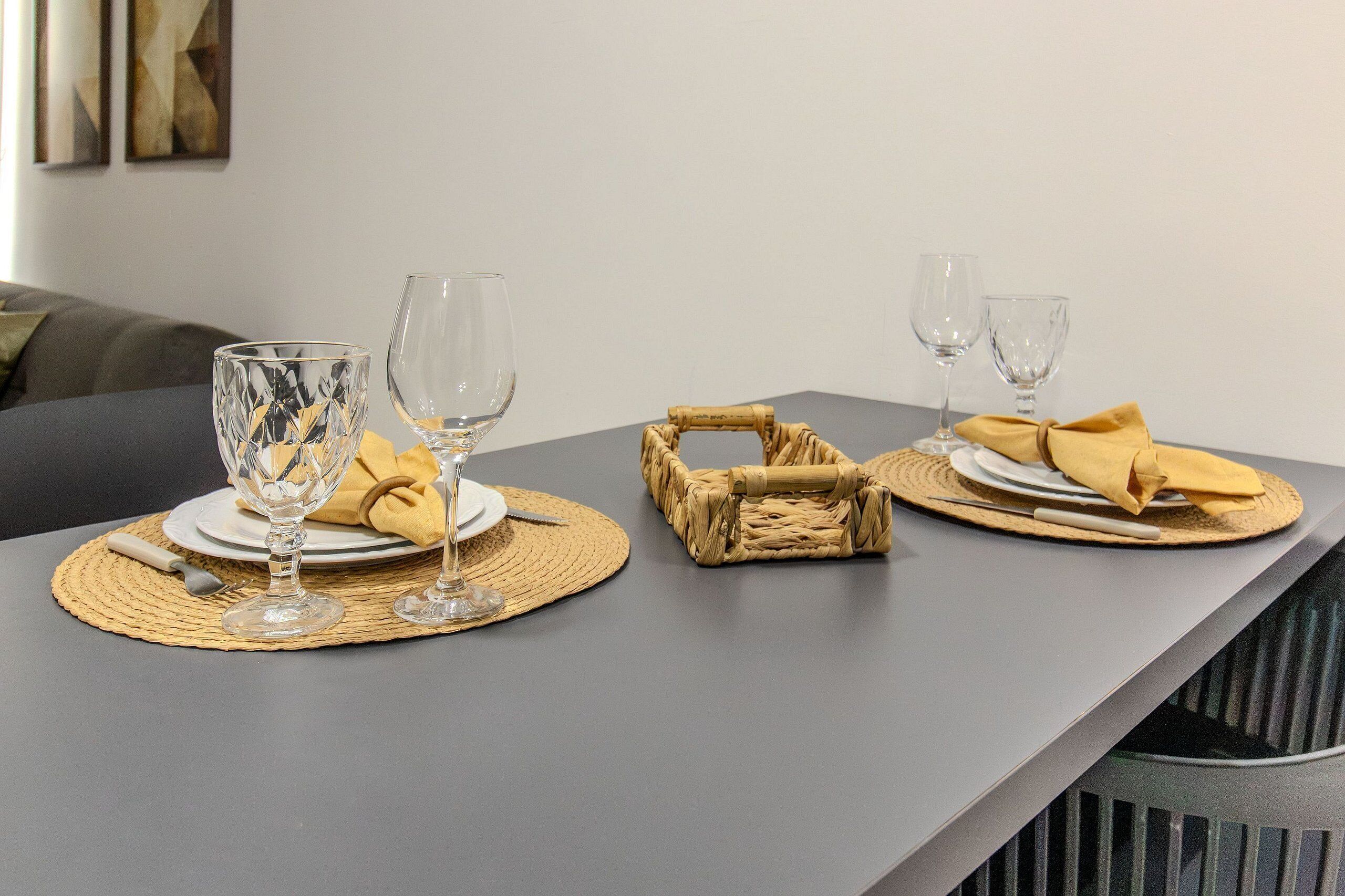 Mesa de jantar posta com dois pratos, duas taças de vinho e uma cesta de fibra.