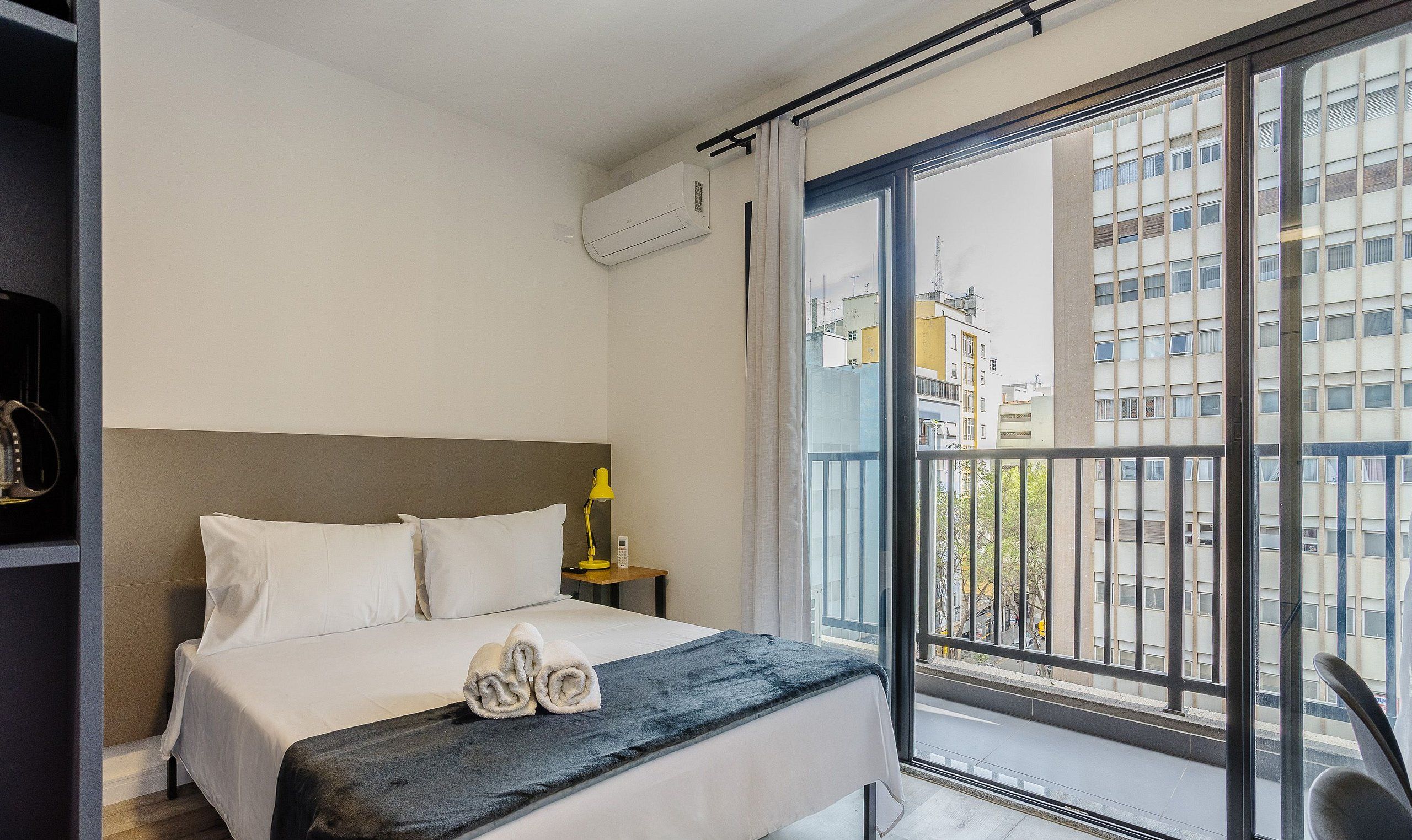 Quarto de hotel com cama, mesa de cabeceira com luminária, ar-condicionado e vista da cidade através de uma janela ampla.