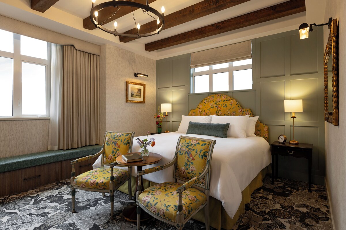 Quarto de hotel com cama, duas poltronas florais, uma mesinha, um banco e uma mesa de cabeceira com abajur.