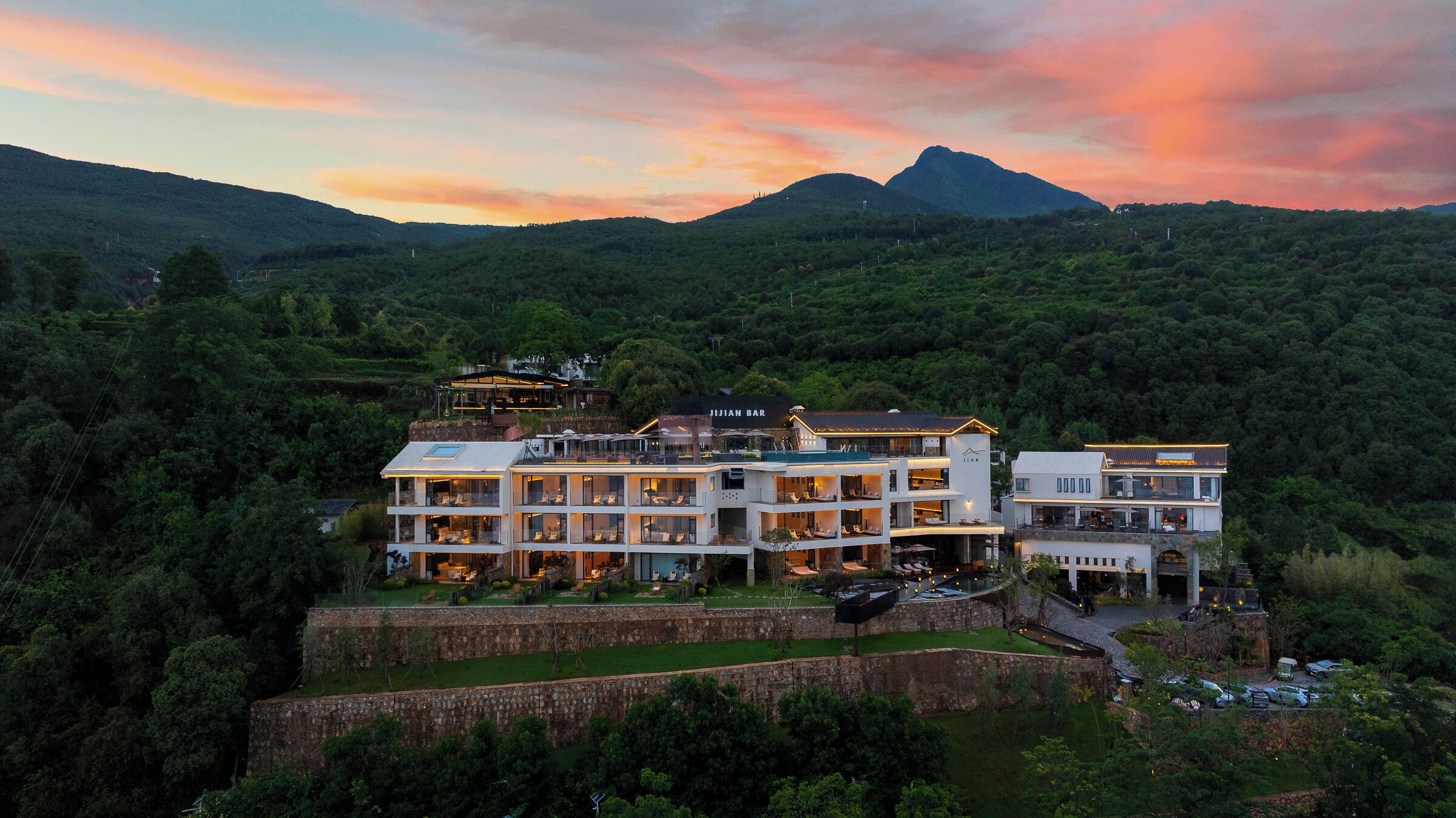 Um hotel de vários andares com um bar no terraço, rodeado por vegetação exuberante e montanhas ao fundo.