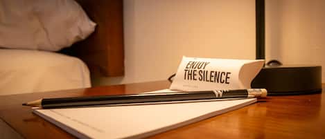 Quarto de hotel com cama, mesa de cabeceira, um livro intitulado 'Enjoy the Silence', um lápis e um objeto preto que pode ser um controle remoto.