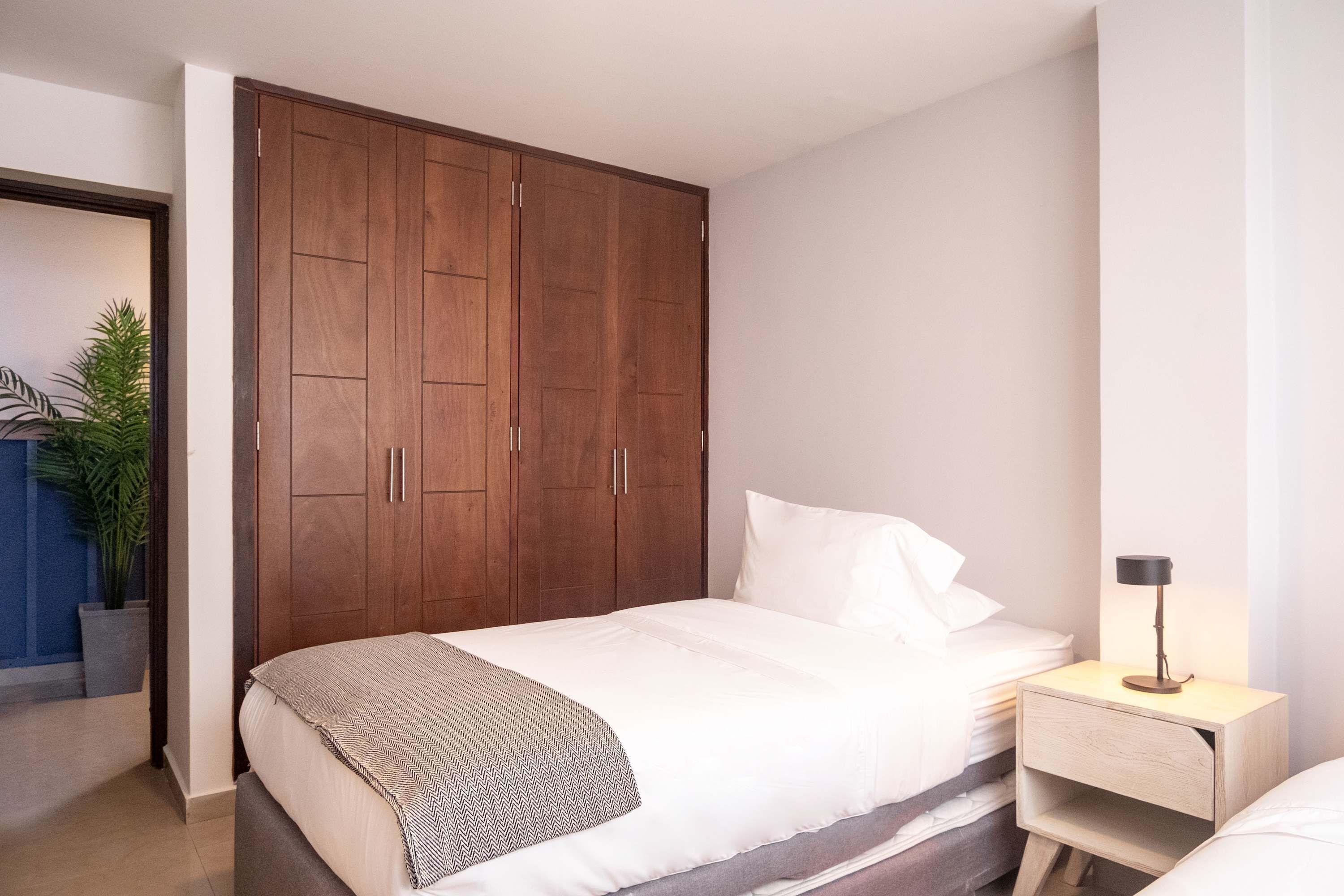 Quarto com um guarda-roupa de madeira grande, uma cama com roupa de cama branca, uma mesa de cabeceira com abajur e uma planta em vaso.