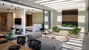 Quarto de hotel moderno com uma cama grande, um sofá, um espaço de refeições e uma pequena varanda com plantas.