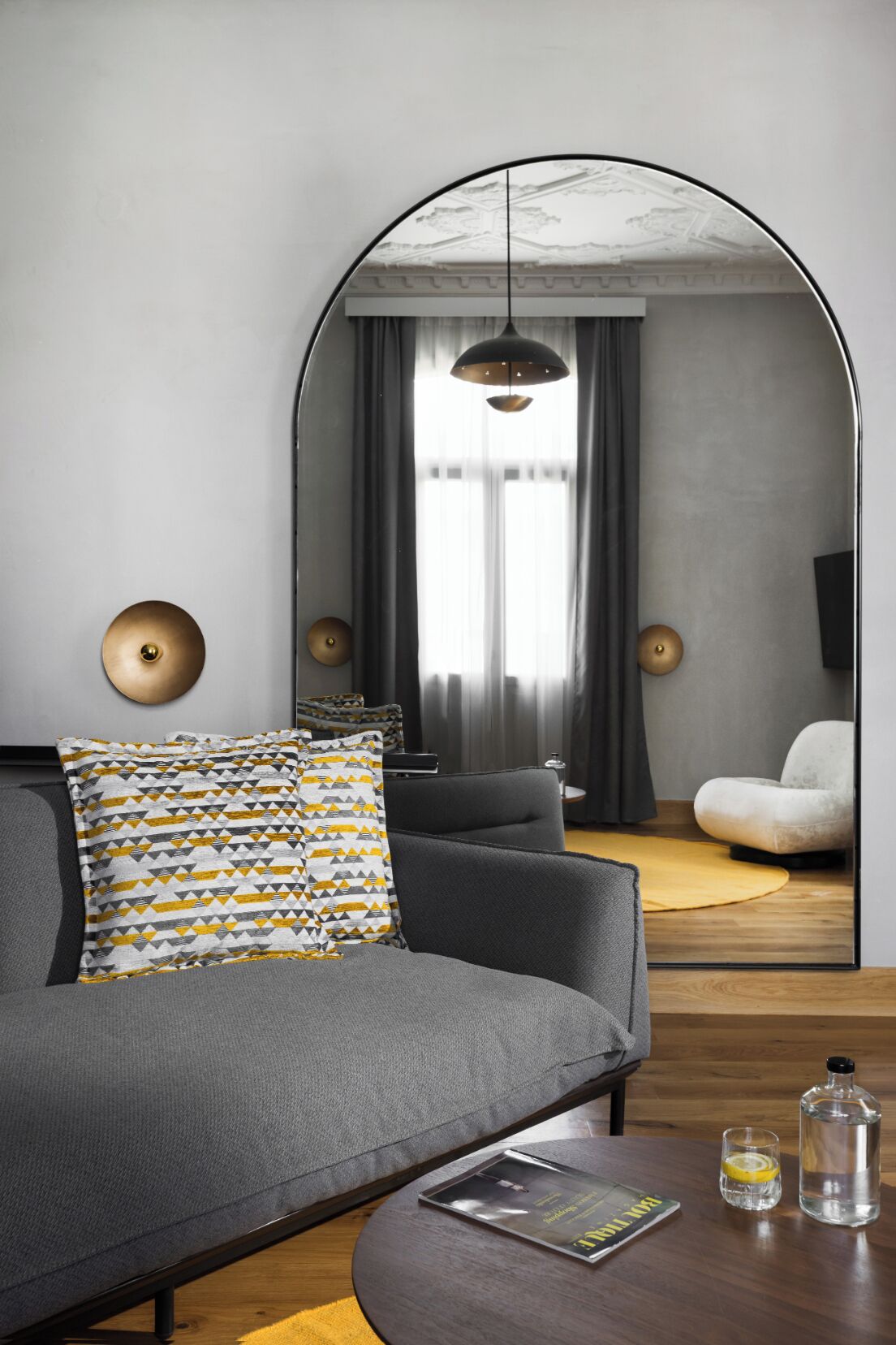 Uma sala de estar moderna com um sofá cinza, almofadas estampadas, uma mesa de centro redonda, um livro, um copo de água e uma garrafa. Um espelho grande reflete o ambiente, e há uma janela com cortinas.