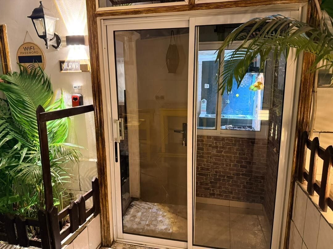 Entrada com porta de vidro para um espaço com piso ladrilhado, planta em vaso e parede de tijolos visíveis através do vidro.