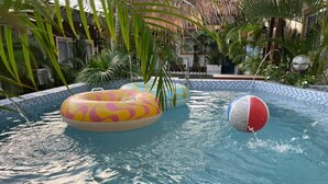 Piscina com boias e uma bola, rodeada por palmeiras e uma borda revestida de azulejos.