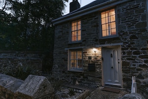 Uma casa de pedra com duas janelas e uma porta, rodeada por um jardim com pedras e plantas.