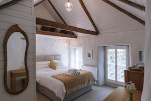 Quarto com cama, cabeceira de madeira, espelho, mesa de cabeceira, cômoda, janela com persianas e luminária de teto.