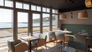 Restaurante com grandes janelas oferecendo vista para o mar e colinas. Mesas montadas com pratos e copos.
