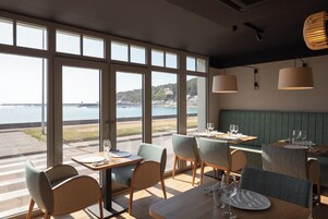 Restaurante com grandes janelas oferecendo vista para o mar e colinas. Mesas montadas com pratos e copos.