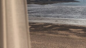 Vista pela janela de uma praia, com montanhas ao longe e céu claro.