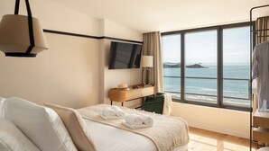 Quarto de hotel com cama, televisão, escrivaninha e uma grande janela com vista para o mar.