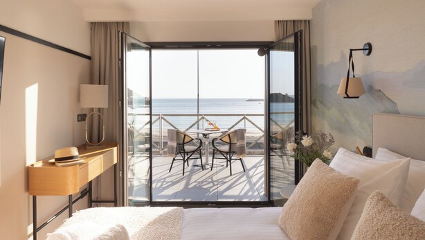 Quarto de hotel com uma cama grande, uma escrivaninha com candeeiro, vista para o mar através de uma porta de vidro e um quadro na parede.