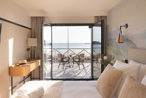 Quarto de hotel com uma cama grande, uma escrivaninha com candeeiro, vista para o mar através de uma porta de vidro e um quadro na parede.