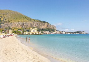 Uma praia com águas turquesa cristalinas, um areal e um edifício com fachada amarela ao fundo.