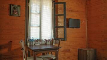 Um quarto com painéis de madeira, uma mesa e cadeiras, uma janela com cortina, uma televisão fixada na parede e um armário pequeno.